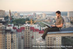 Man perched on roof with legs dangling over city while working on ...